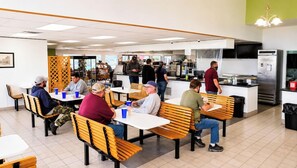 Uma cafeteria com mesas, bancos, área de cozinha e balcão onde pessoas estão pedindo comida.