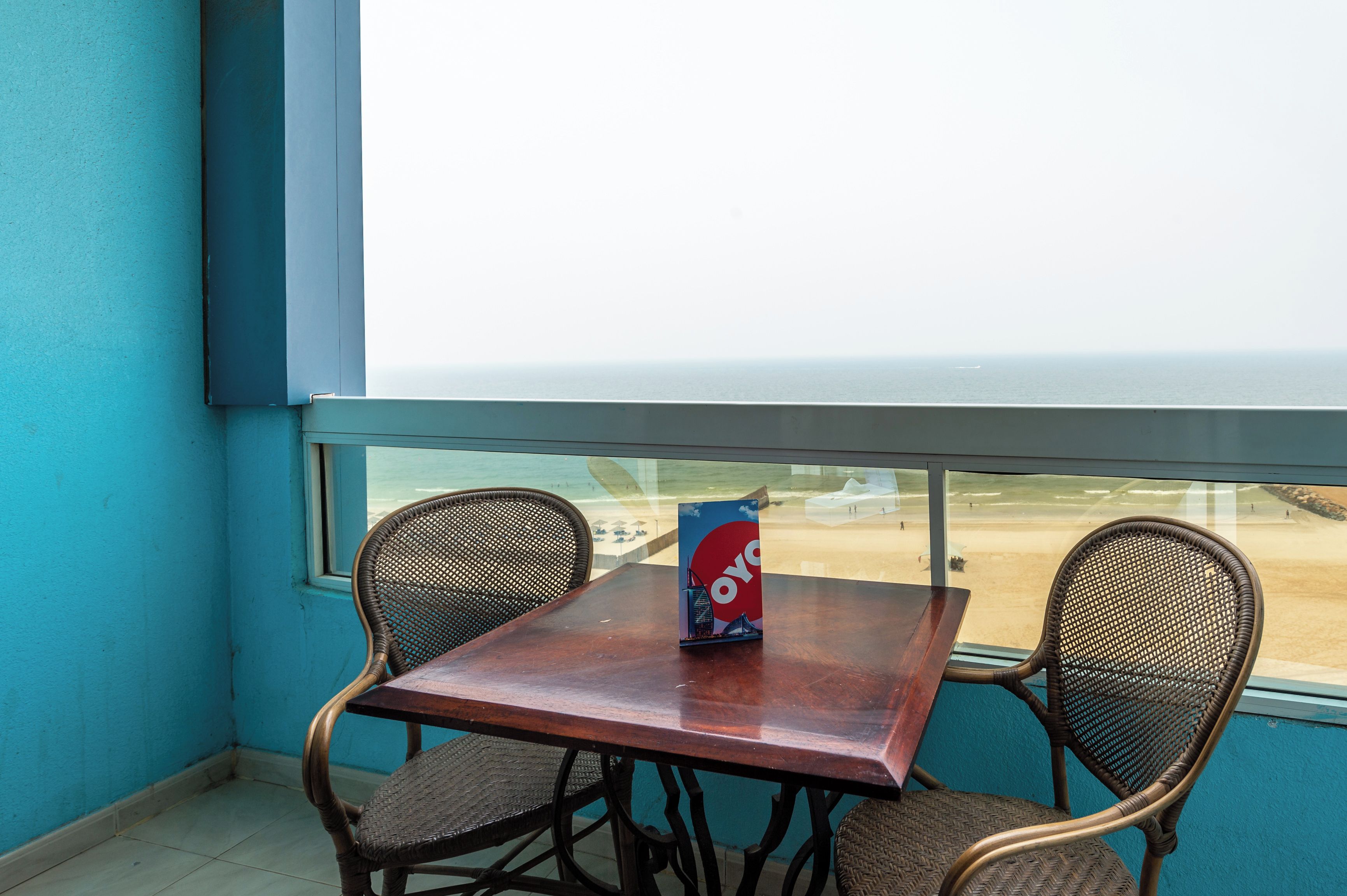 Uma mesa com um saco de snacks OYO, duas cadeiras de vime e vista para a praia através de uma janela.