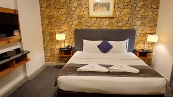 Quarto de hotel com parede de pedra, televisão de tela plana, cama com cabeceira cinza, duas luminárias de cabeceira e um quadro na parede.