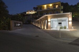 Um hotel moderno com entrada bem iluminada, uma placa visível e um edifício de vários andares ao fundo.