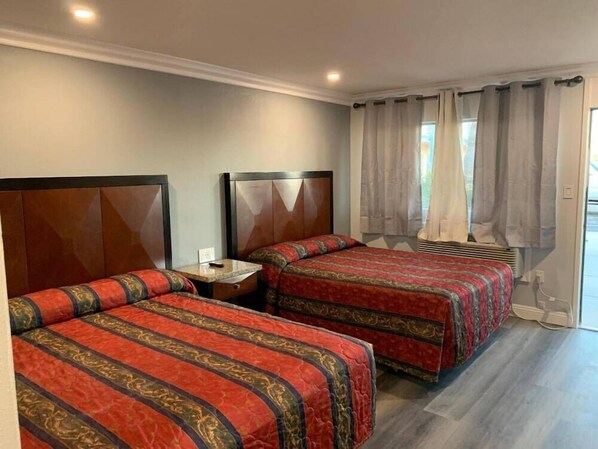 Quarto de hotel com duas camas, uma pequena mesa de cabeceira, uma janela com cortinas e uma lâmpada no teto.