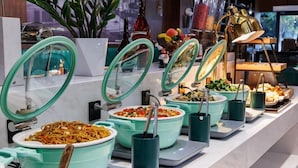 Buffet com diversos pratos em recipientes de cor verde-água, uma balança para pesar alimentos e um abajur no lado direito.