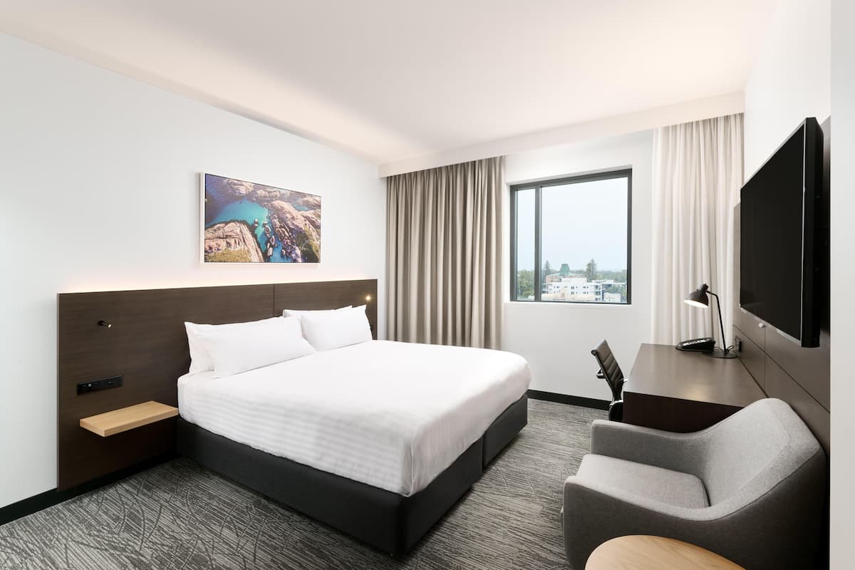 Quarto de hotel com uma cama, escrivaninha, cadeira, televisão, janela com cortinas e um quadro na parede.