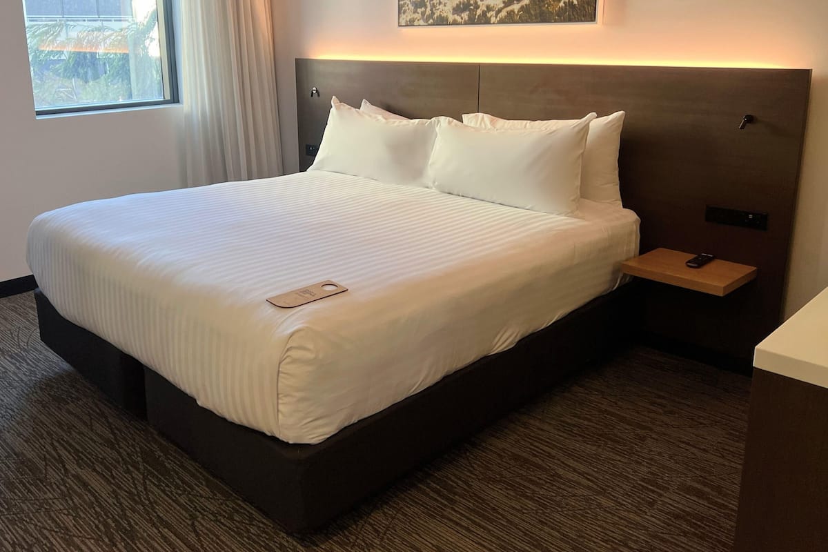 Quarto de hotel com uma cama grande, mesa de cabeceira com controle remoto, criado-mudo e uma janela com cortinas.