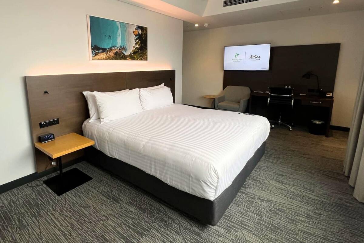 Quarto de hotel com uma cama grande, mesa de cabeceira com relógio, escrivaninha com cadeira e uma tela de TV exibindo o nome do hotel.
