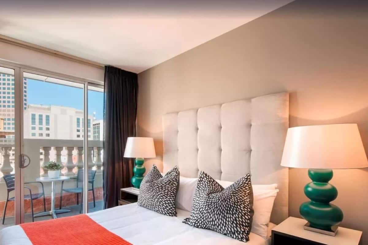 Quarto de hotel com uma cama grande, duas luminárias de cabeceira, uma varanda com vista para edifícios e um cobertor vermelho.