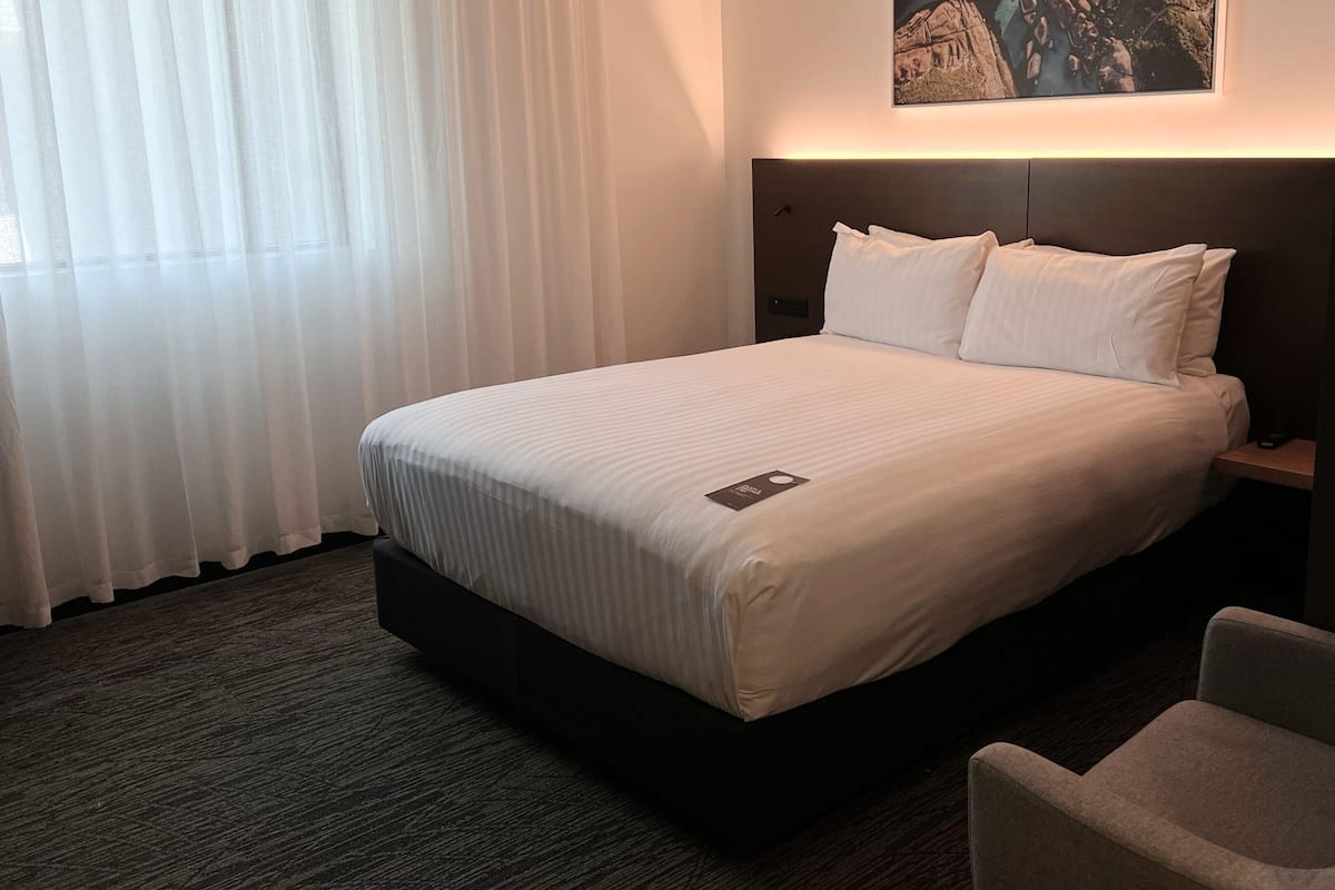 Quarto de hotel com cama, mesa de cabeceira, cadeira, pintura na parede e janela com cortinas.