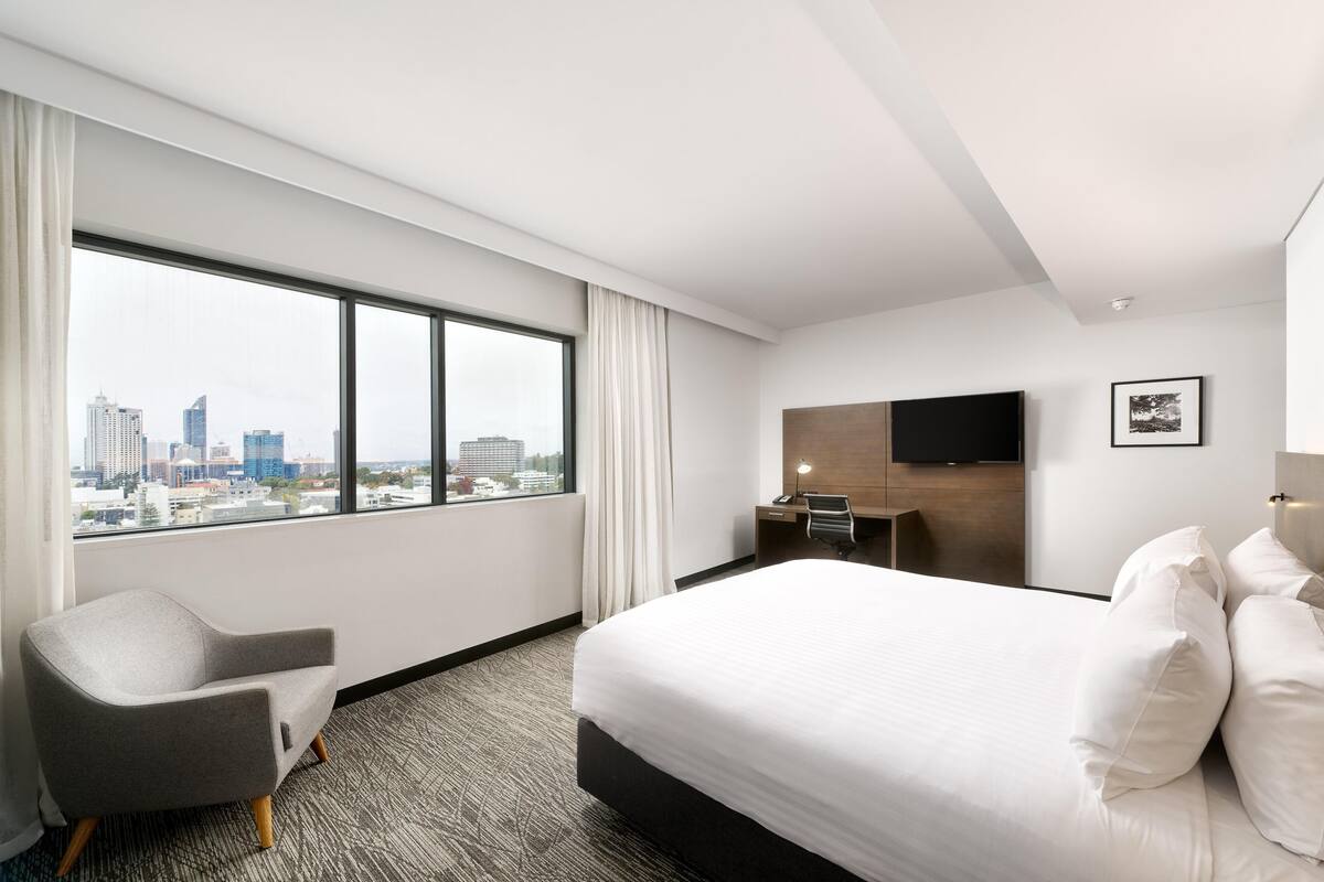 Quarto de hotel com uma grande janela, cama, cadeira, escrivaninha e TV de tela plana.