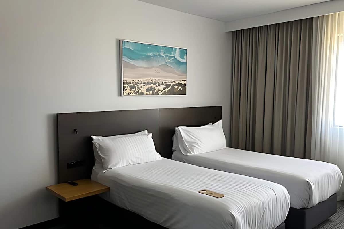 Quarto de hotel com duas camas, uma mesa de cabeceira, um controle remoto, uma pintura de paisagem desértica e uma janela grande com cortinas.