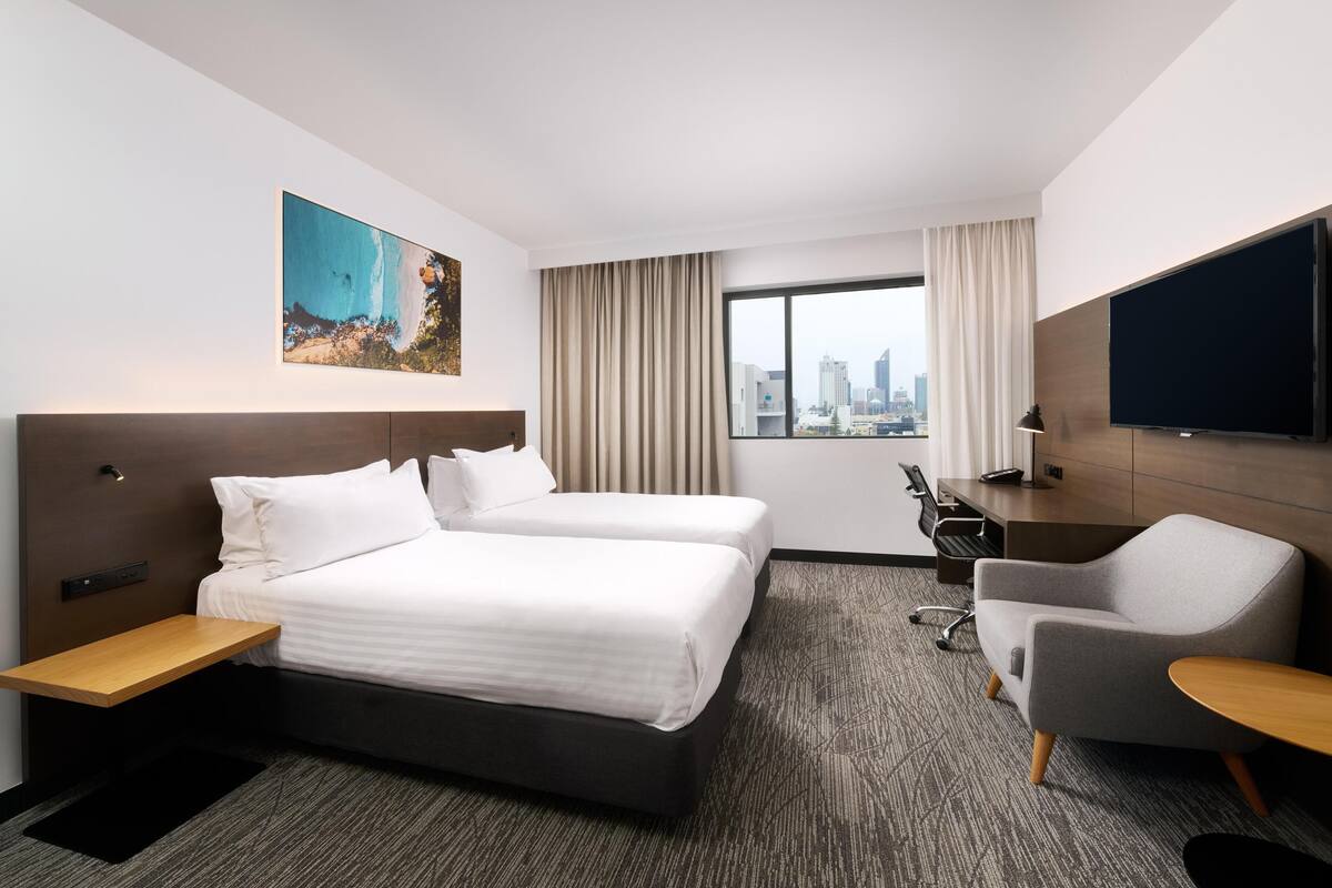 Quarto de hotel com uma cama, duas mesas de cabeceira, uma escrivaninha, uma cadeira, televisão e uma janela grande com vista para a cidade.