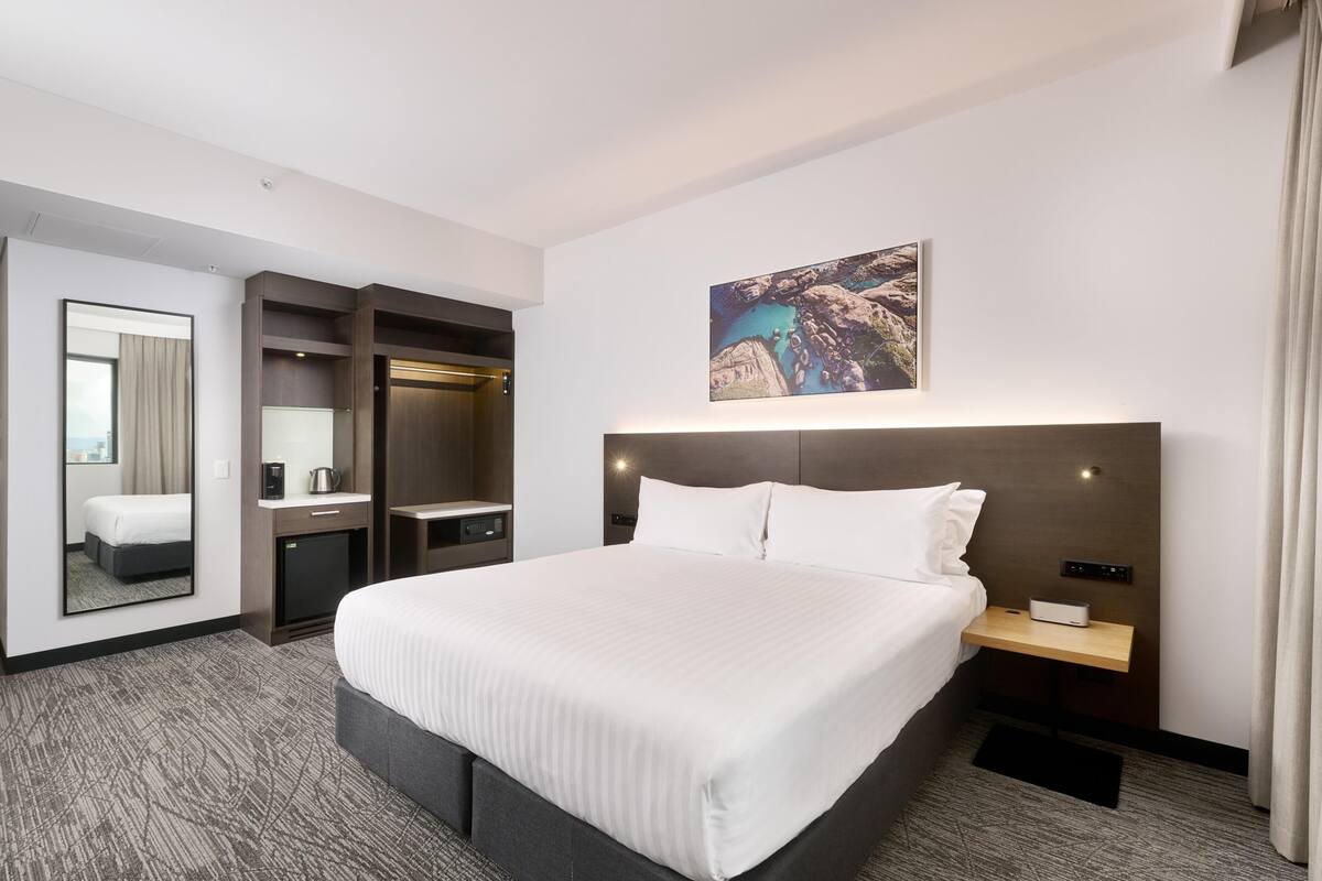 Quarto de hotel com cama grande, mesa de cabeceira, espelho, guarda-roupa e um quadro na parede.
