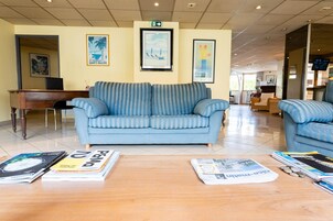 Um hall com um sofá azul, uma mesa de centro de madeira com revistas, um piano e quadros nas paredes.