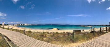 Uma praia com um calçadão de madeira que leva até a costa, águas turquesa cristalinas e alguns edifícios ao fundo.