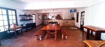 Uma ampla área de refeições com uma grande mesa de madeira e bancos, uma cozinha com bancada de pedra e uma televisão fixada na parede.