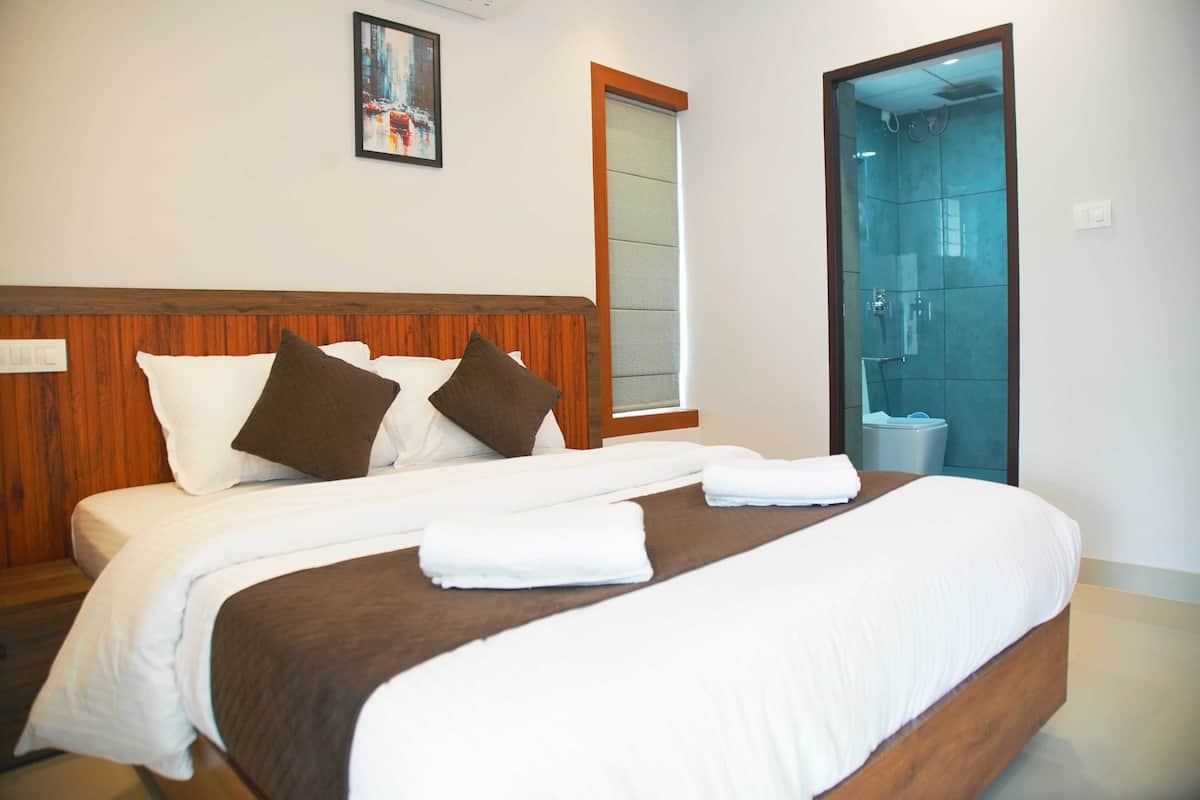 Quarto de hotel com cama grande, cabeceira de madeira, roupa de cama branca e marrom, quadro na parede e porta de box de vidro.