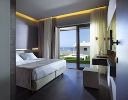 Quarto de hotel moderno com uma cama grande, mesa de cabeceira pequena, espelho e vista para o mar através de uma porta de vidro deslizante.