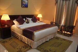 Quarto de hotel com uma cama grande, duas luminárias de cabeceira, uma cadeira, uma mesinha e um quadro na parede.