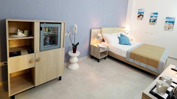 Quarto de hotel com cama, mesa de cabeceira com abajur, pequena mesa branca com um vaso de flores, guarda-roupa e uma prateleira com diversos objetos.