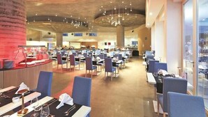 Um restaurante amplo com várias mesas e cadeiras, teto alto com luzes pendentes e vista através de grandes janelas.
