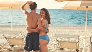 Um casal se abraçando na praia, com guarda-sóis e cadeiras de praia.
