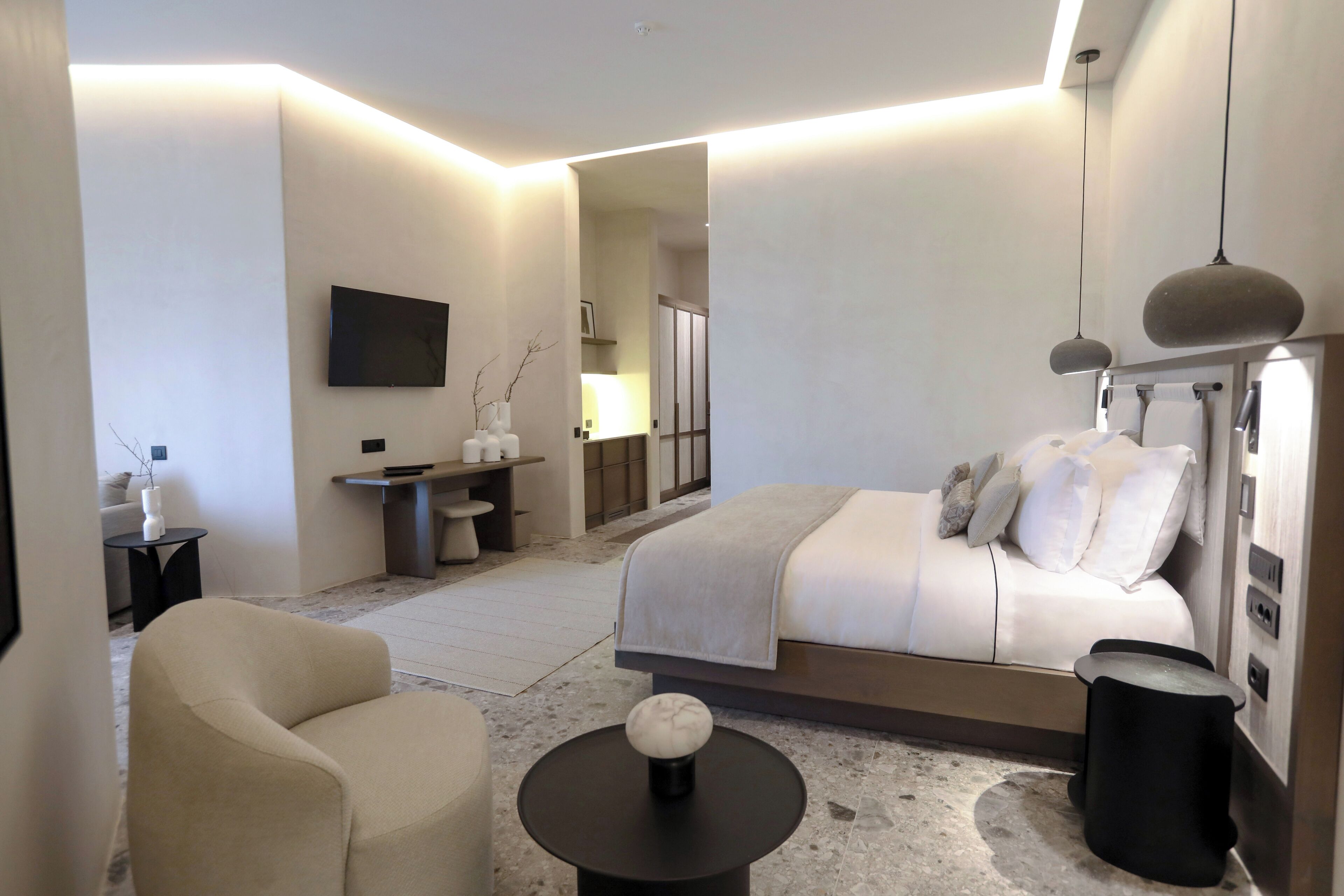 Quarto de hotel moderno com cama, mesa de cabeceira, cadeira e uma mesa redonda com um objeto decorativo.