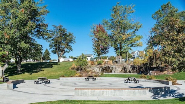 Um parque com mesas de piquenique, um espaço de estar circular central e árvores ao redor.