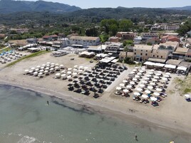 Vista aérea de uma praia com fileiras de espreguiçadeiras e guarda-sóis, um edifício com teto plano e montanhas ao fundo.