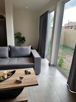 Uma sala de estar moderna com um sofá cinzento, uma mesa de centro de madeira e uma janela grande com cortinas.