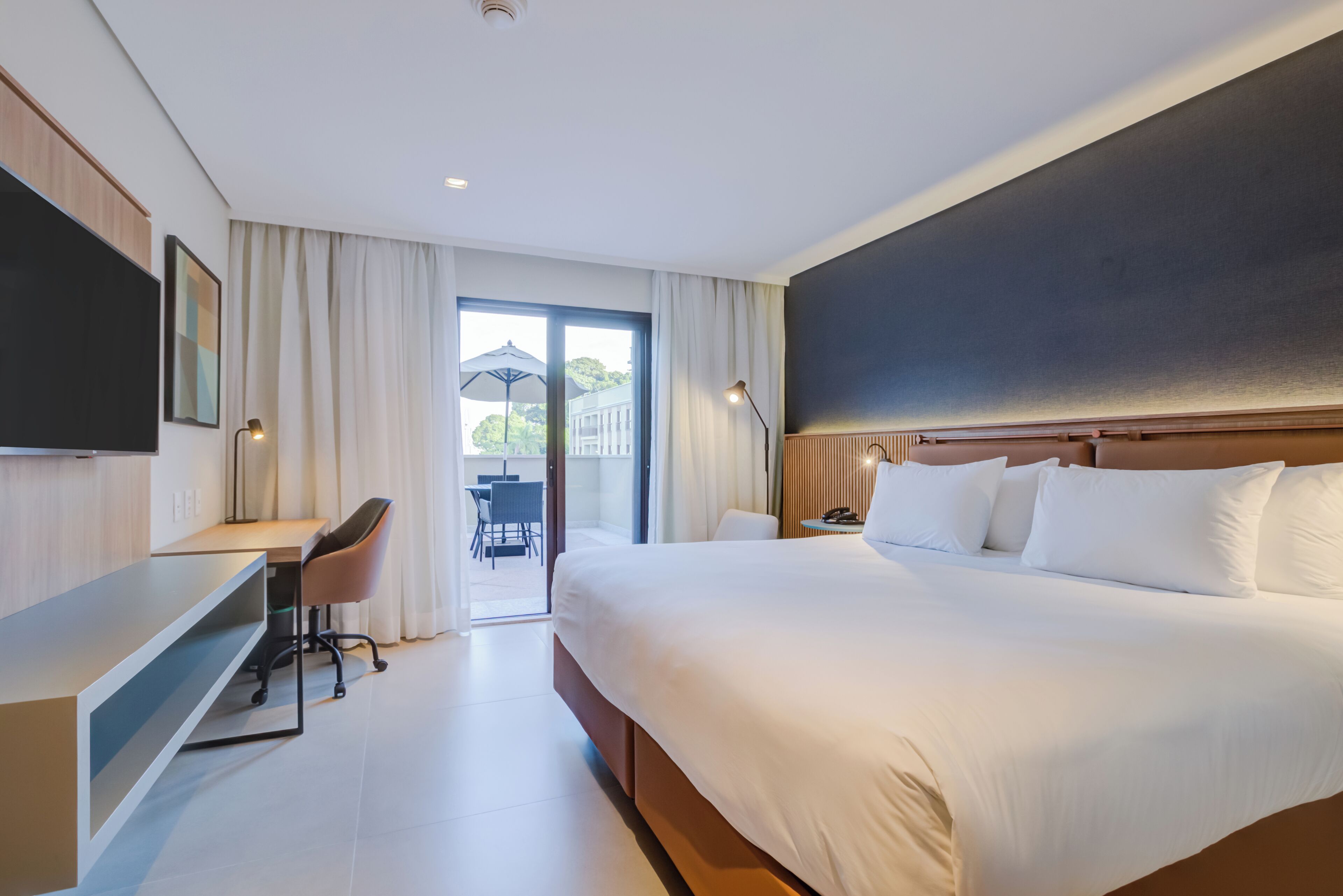 Quarto de hotel moderno com uma cama grande, uma escrivaninha, uma cadeira, uma televisão e uma varanda com guarda-sol.