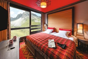 Quarto de hotel com cama, escrivaninha com cadeira, janela e vista para montanhas, além de colcha xadrez vermelha e branca.