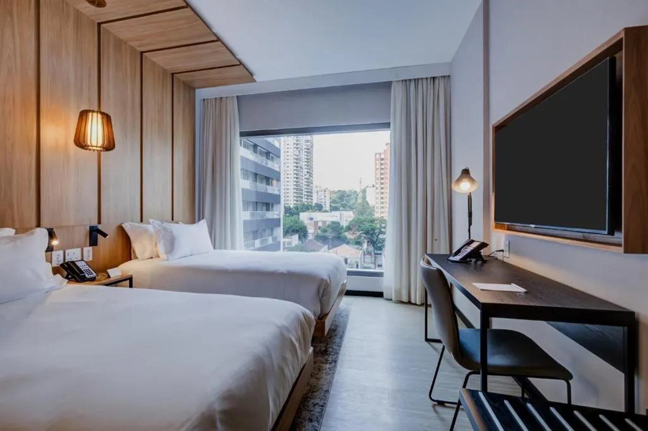 Quarto de hotel com duas camas, uma escrivaninha, uma cadeira, televisão e janela com vista para a cidade.