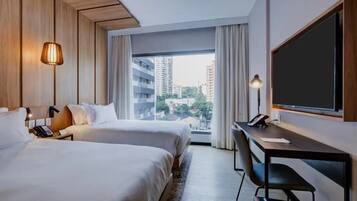 Quarto de hotel com duas camas, uma escrivaninha, uma cadeira, televisão e janela com vista para a cidade.