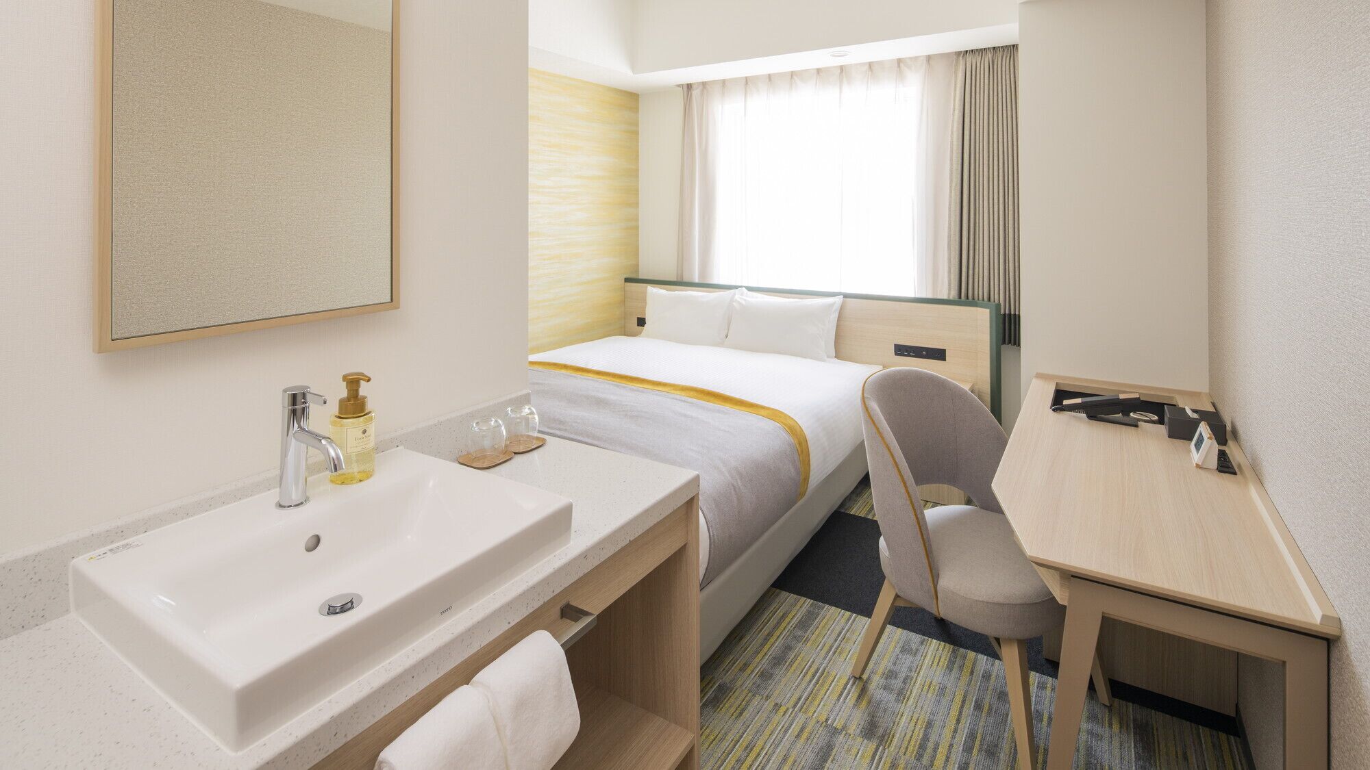 Quarto de hotel com cama, escrivaninha, cadeira, pia e espelho.
