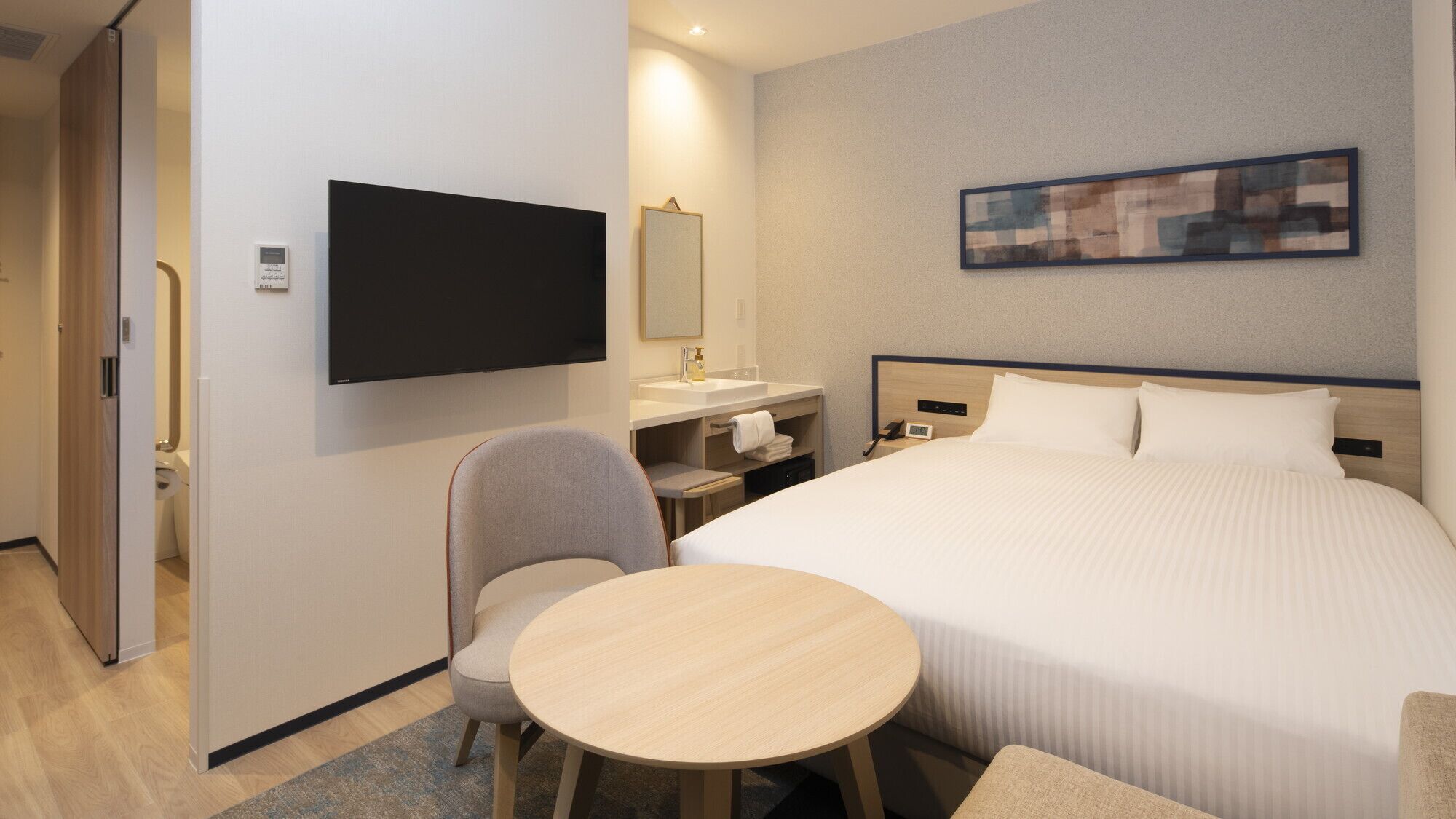 Quarto de hotel com cama, escrivaninha, cadeira, televisão, banheiro e um quadro na parede.