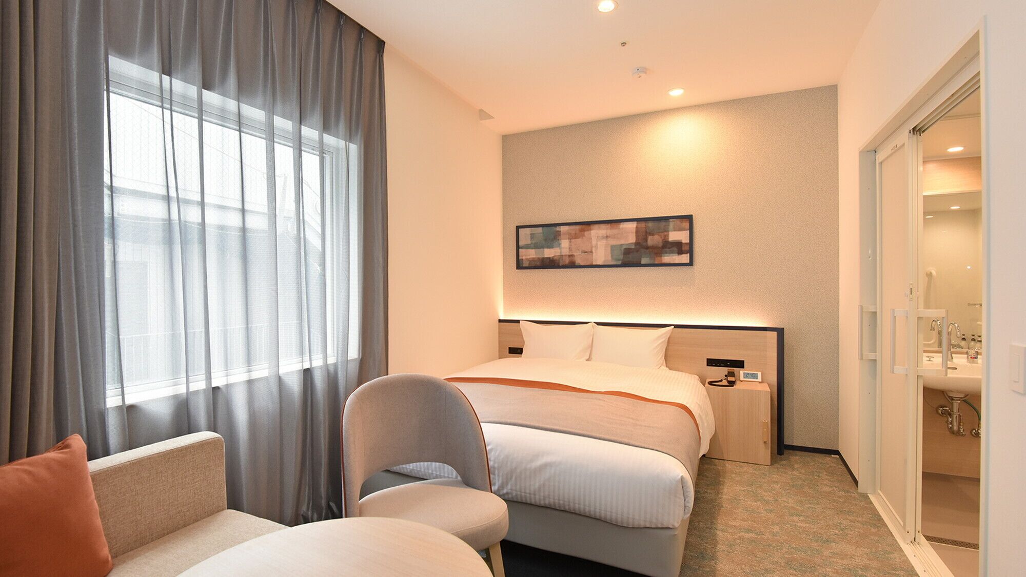 Quarto de hotel com cama, uma cadeira, mesa de cabeceira, quadro na parede e banheiro visível por uma porta aberta.
