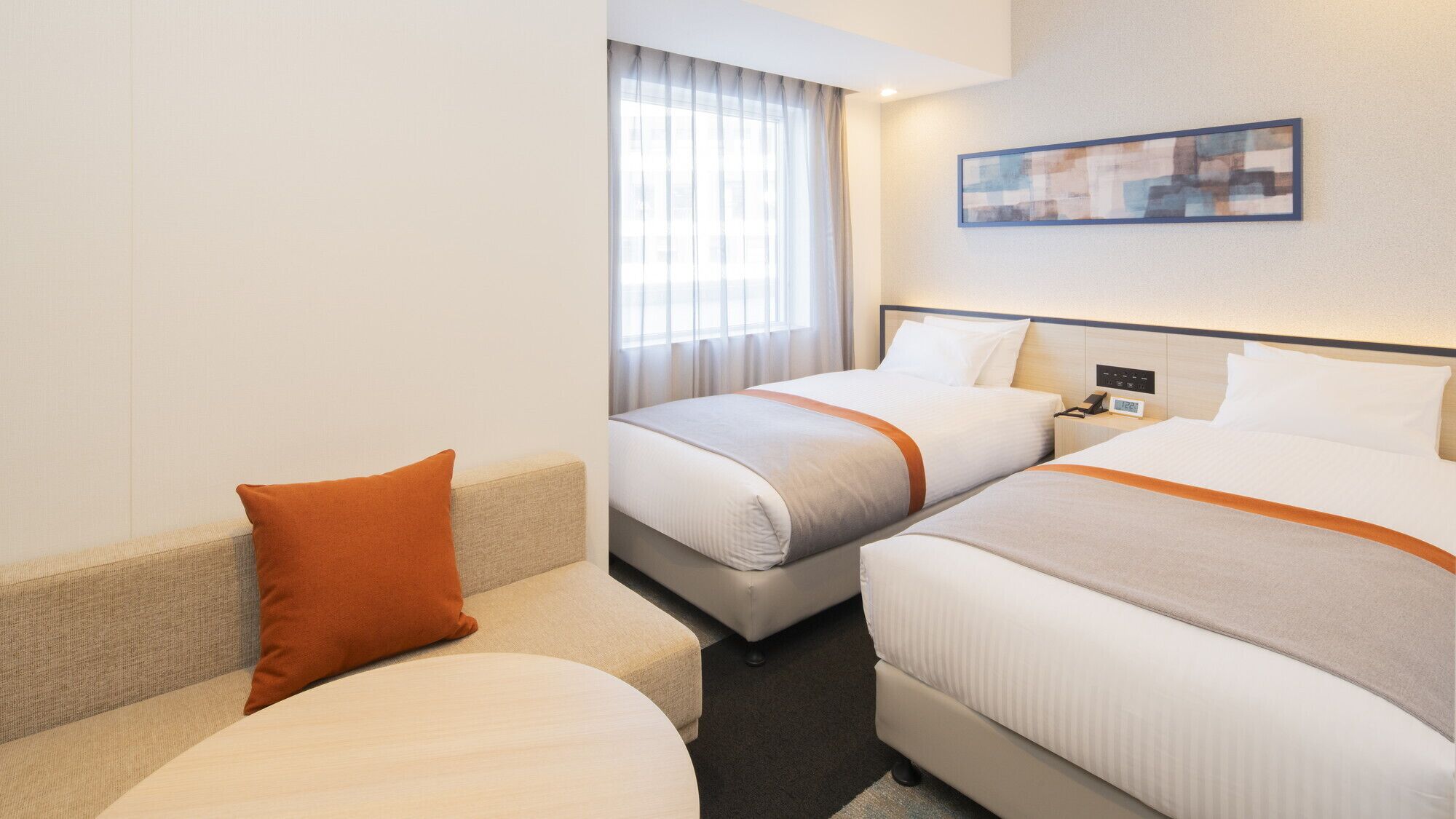 Quarto de hotel com duas camas, um sofá, janela com cortinas, um quadro na parede e mesa de cabeceira com telefone e relógio.