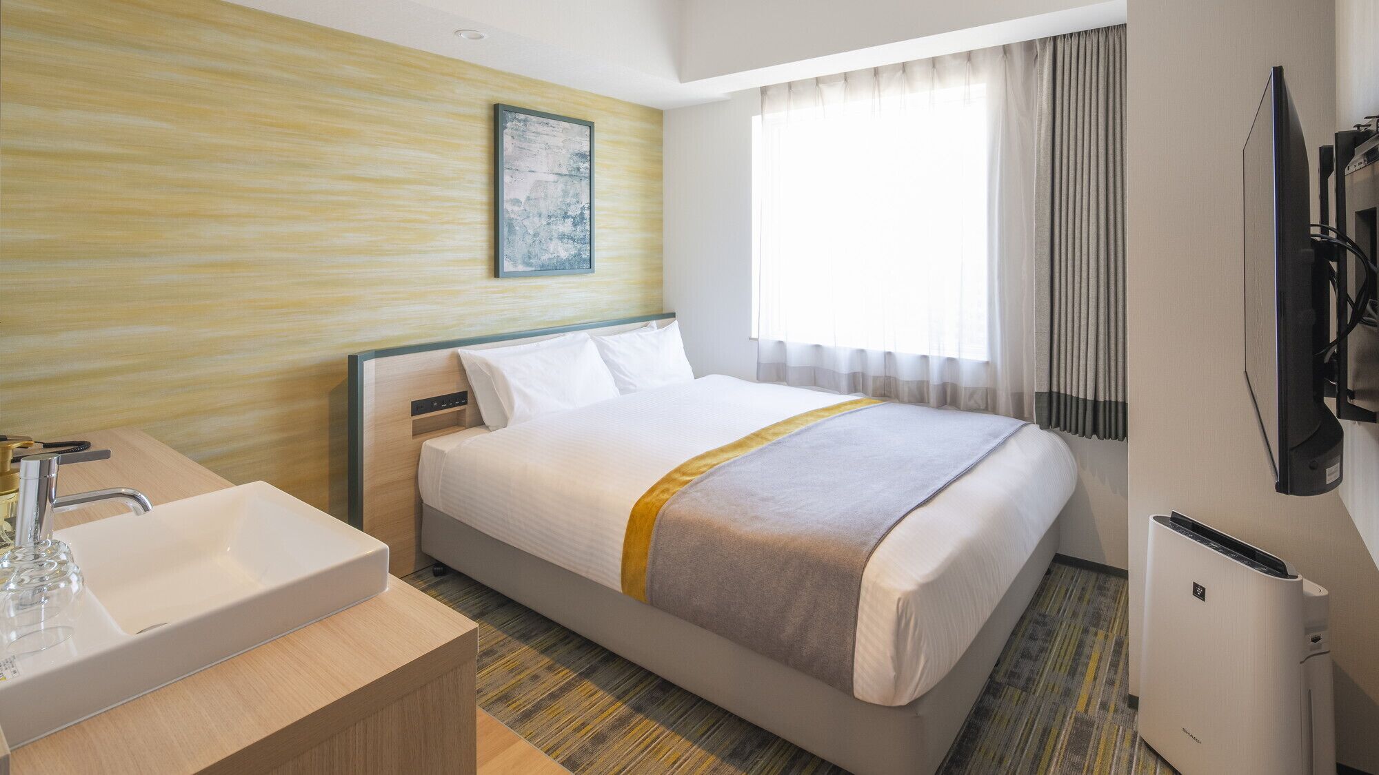 Quarto de hotel com cama, mesa de cabeceira, pia, televisão e janela com cortinas.