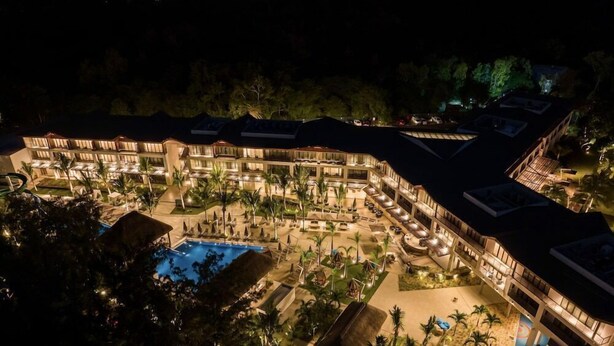 Um resort com vários edifícios, uma piscina central e palmeiras ao redor.