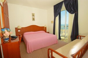 Quarto de hotel com cama, mesa de cabeceira com abajur, uma cadeira e vista para o mar por uma grande janela.