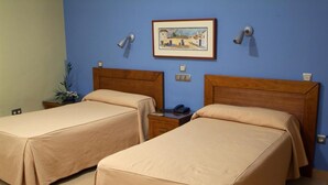 Quarto de hotel com duas camas de solteiro, mesa de cabeceira com abajur, quadro na parede e pequena mesa com um vaso de flores.