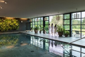Piscina interna com grandes janelas, plantas em vasos e vista para o verde lá fora.