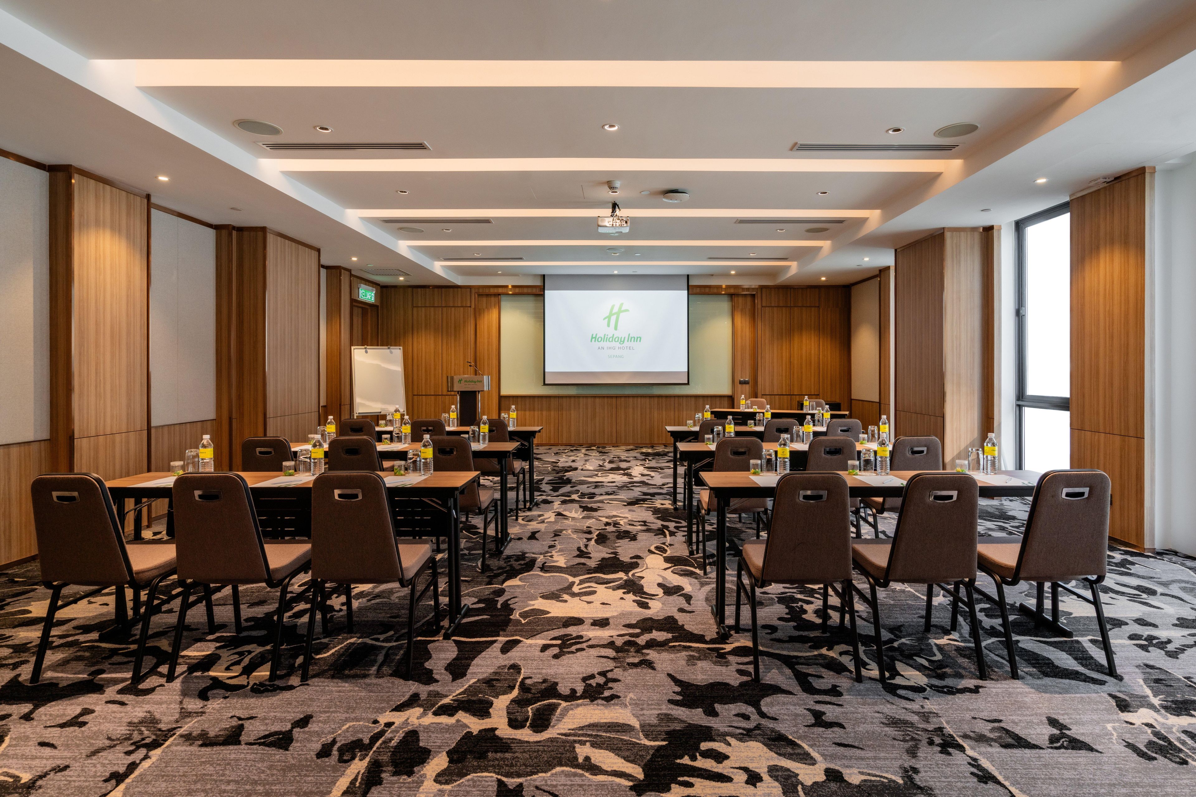 Uma sala de conferências com uma tela grande exibindo a marca 'Holiday Inn', várias mesas com cadeiras e piso coberto de carpete.