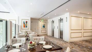 Um espaço de jantar de hotel com mesas preparadas para quatro pessoas, cadeiras e obras de arte nas paredes.
