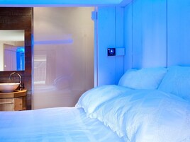 Quarto com cama, mesa de cabeceira, espelho e painel de controle na parede.