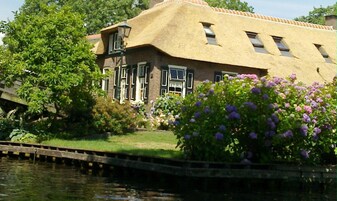 Uma casa com telhado de palha, um jardim com flores roxas e um canal em primeiro plano.