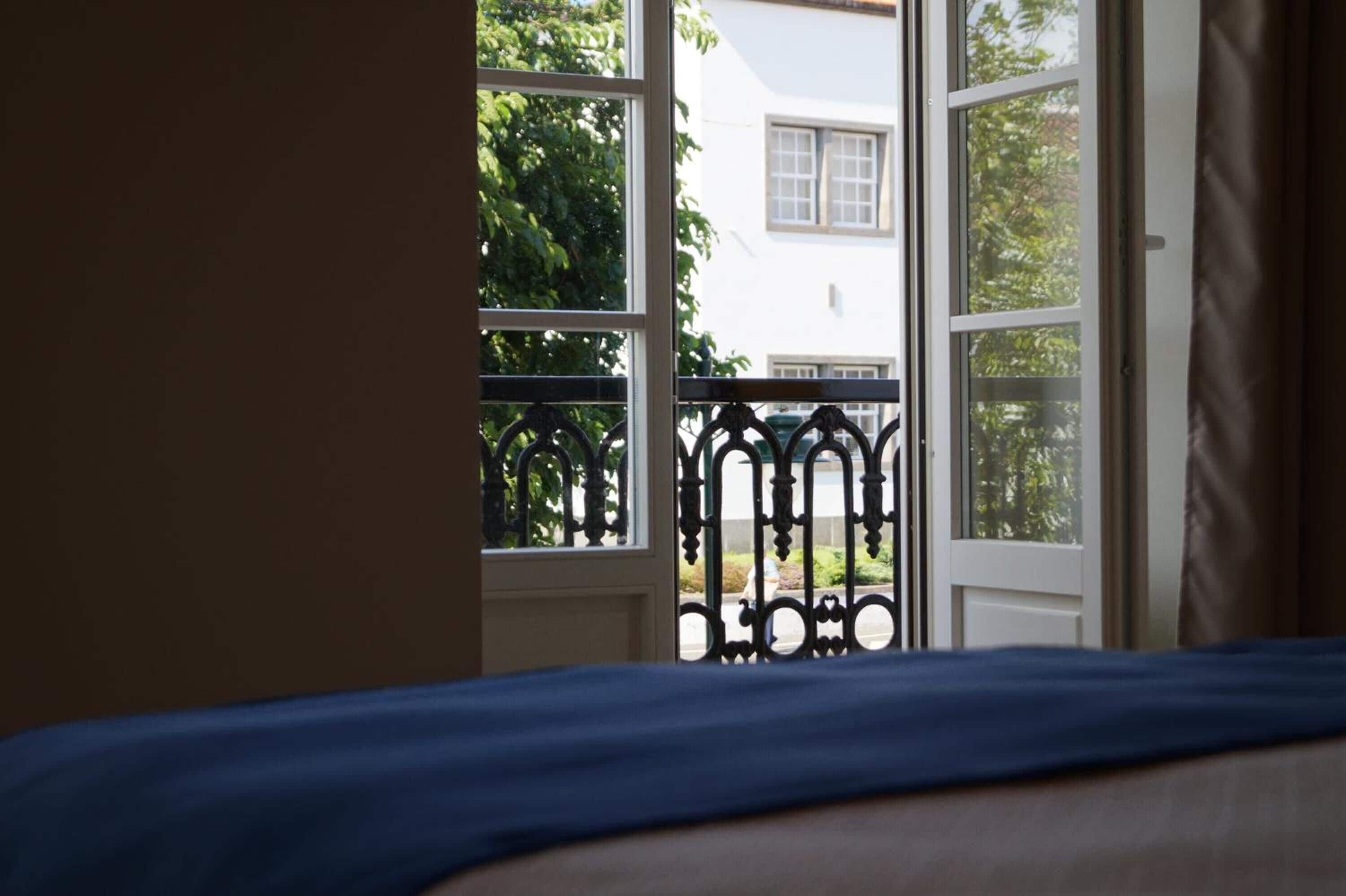 Quarto com cama, janela com grade de metal e vista para um edifício branco.
