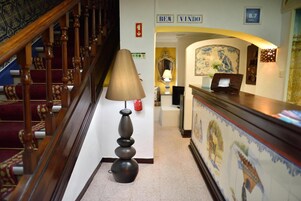 Um saguão de hotel com uma mesa de recepção, uma escadaria com poltronas estofadas vermelhas e um detector de incêndio fixado na parede.