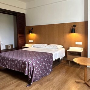 Quarto de hotel com cama grande, duas mesas de cabeceira, uma mesinha redonda e duas luminárias fixadas na parede.