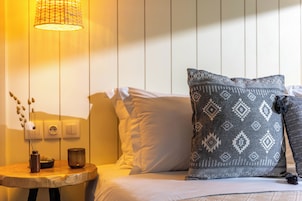 Uma cama bem arrumada com almofadas estampadas, uma pequena mesa de cabeceira de madeira com um vaso e uma vela, e um luminário com luz acolhedora.