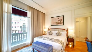 Quarto de hotel com uma cama grande, uma janela com cortinas, uma mesa de cabeceira com abajur e um quadro na parede.